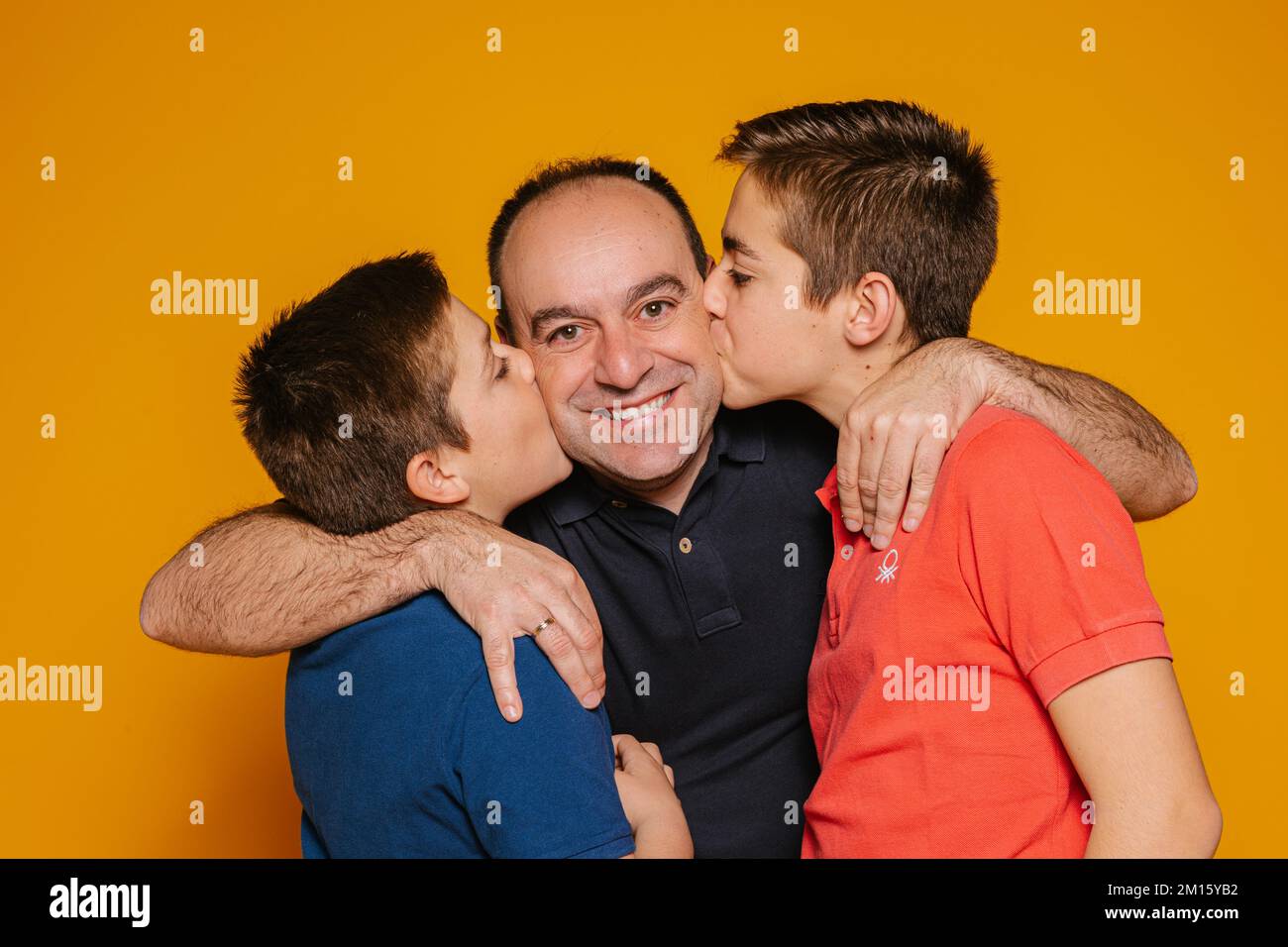 Cheerful sons kissing their single father while standing against yellow ...