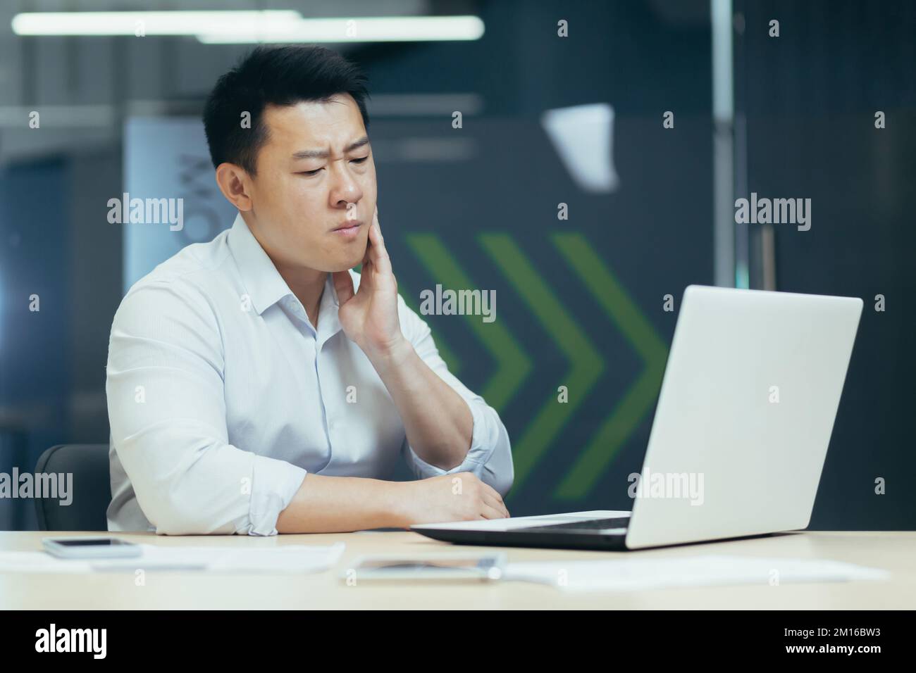 Toothache at work. A young Asian man sits in the office at the table ...