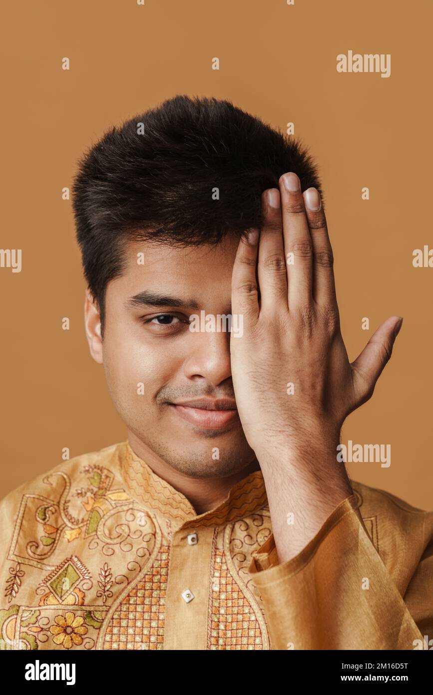 Young brunette indian man smiling and covering his face with hand isolated over yellow ...