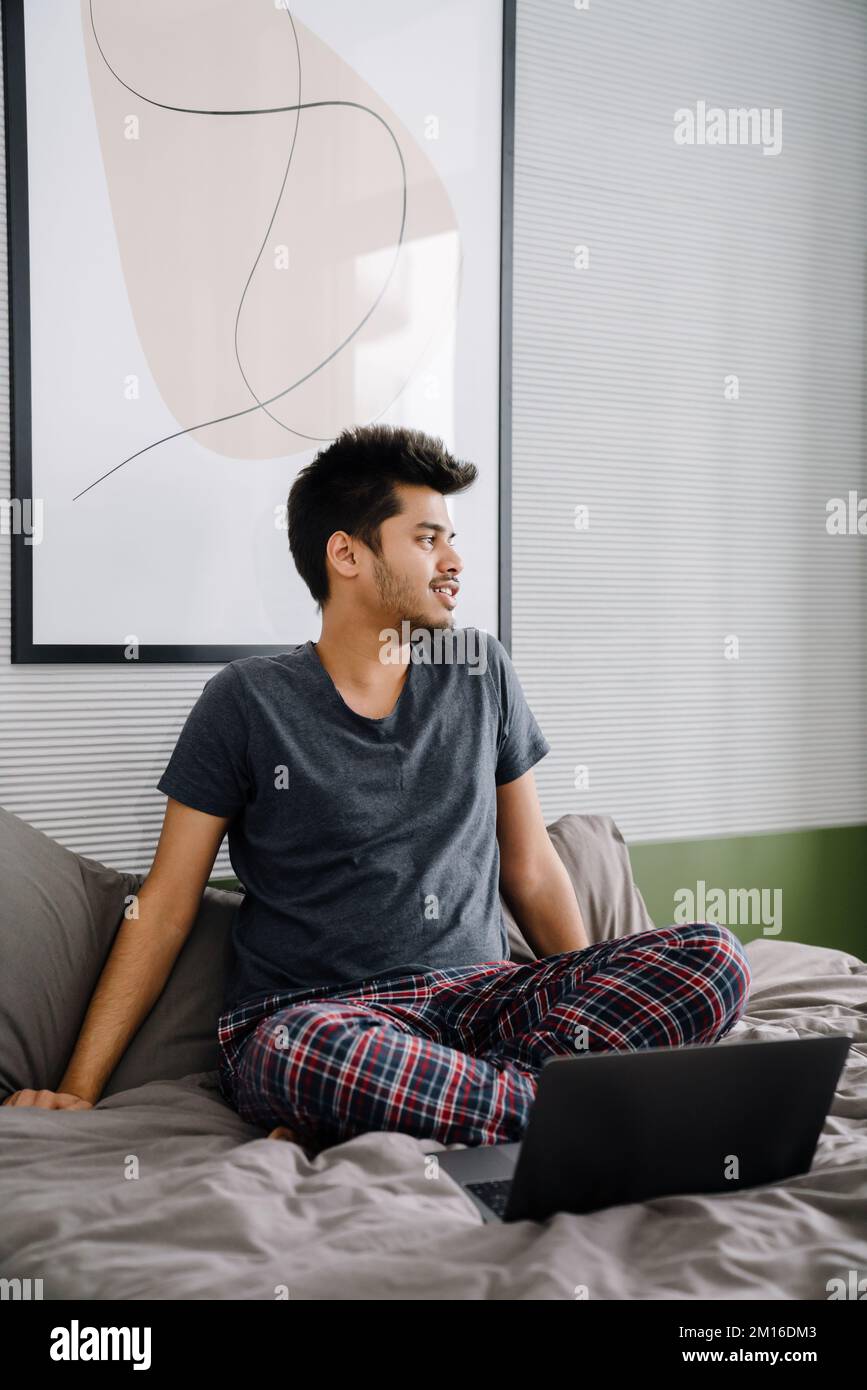 Young handsome indian man sitting on bed in lotus pose with laptop and ...