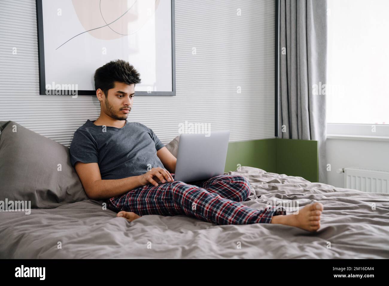 Young handsome indian man liying on bed and working on laptop in cozy ...