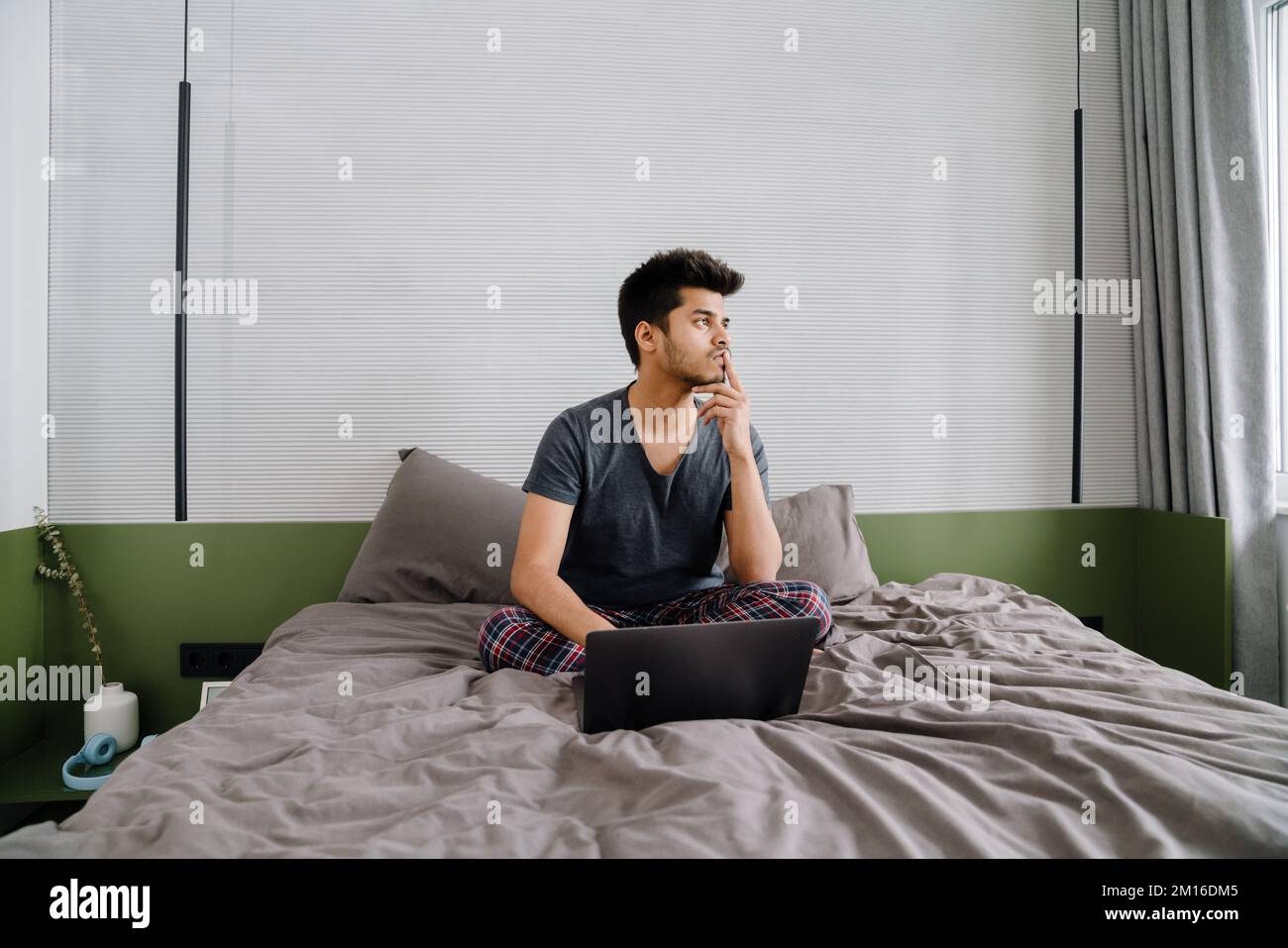 Young handsome thoughtful indian man sitting on bed in lotus pose with ...