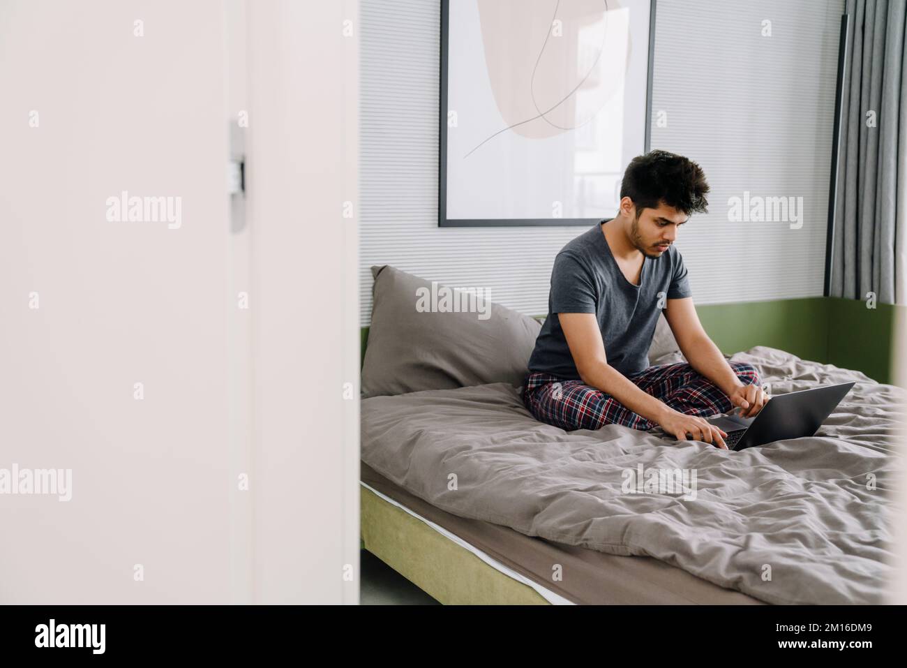 Young handsome indian man sitting on bed and working on laptop in cozy ...