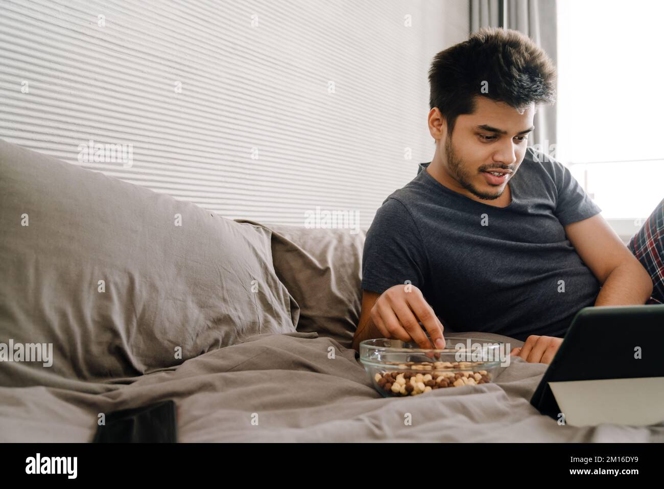Young handsome smiling indian man lying on bed with tablet eating snack ...