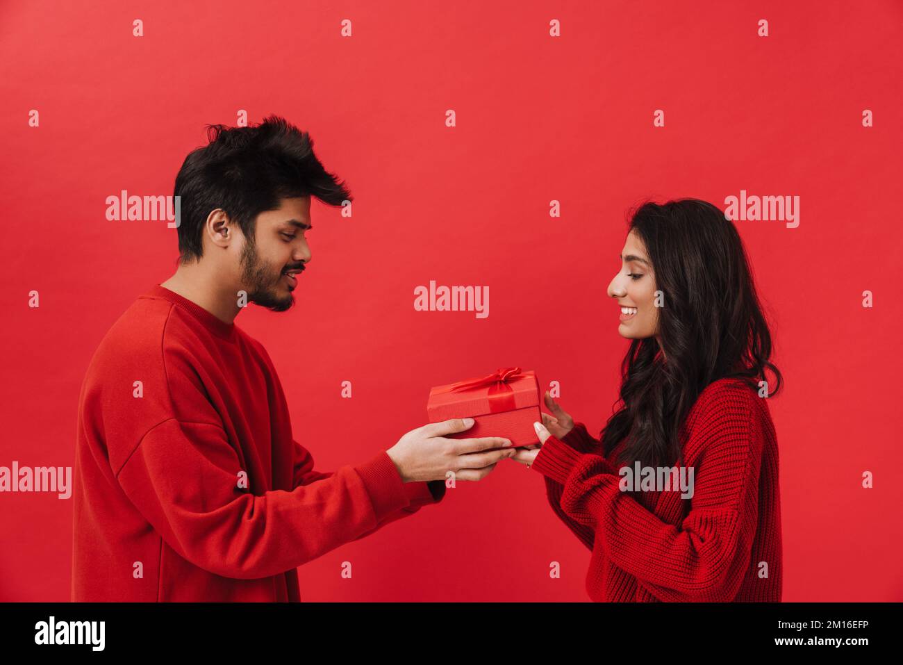 Young indian man giving present box to his girlfriend isolated over red ...