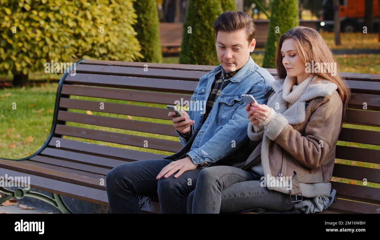Caucasian couple two young people partners sit on bench use phone cell ...