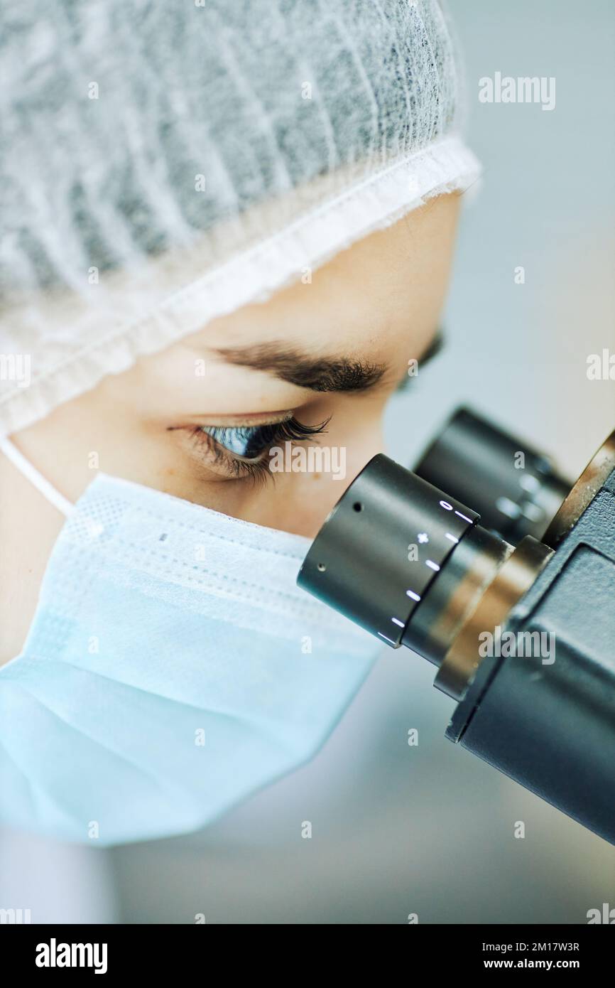 Vertical closeup of young female medic looking in microscope while ...