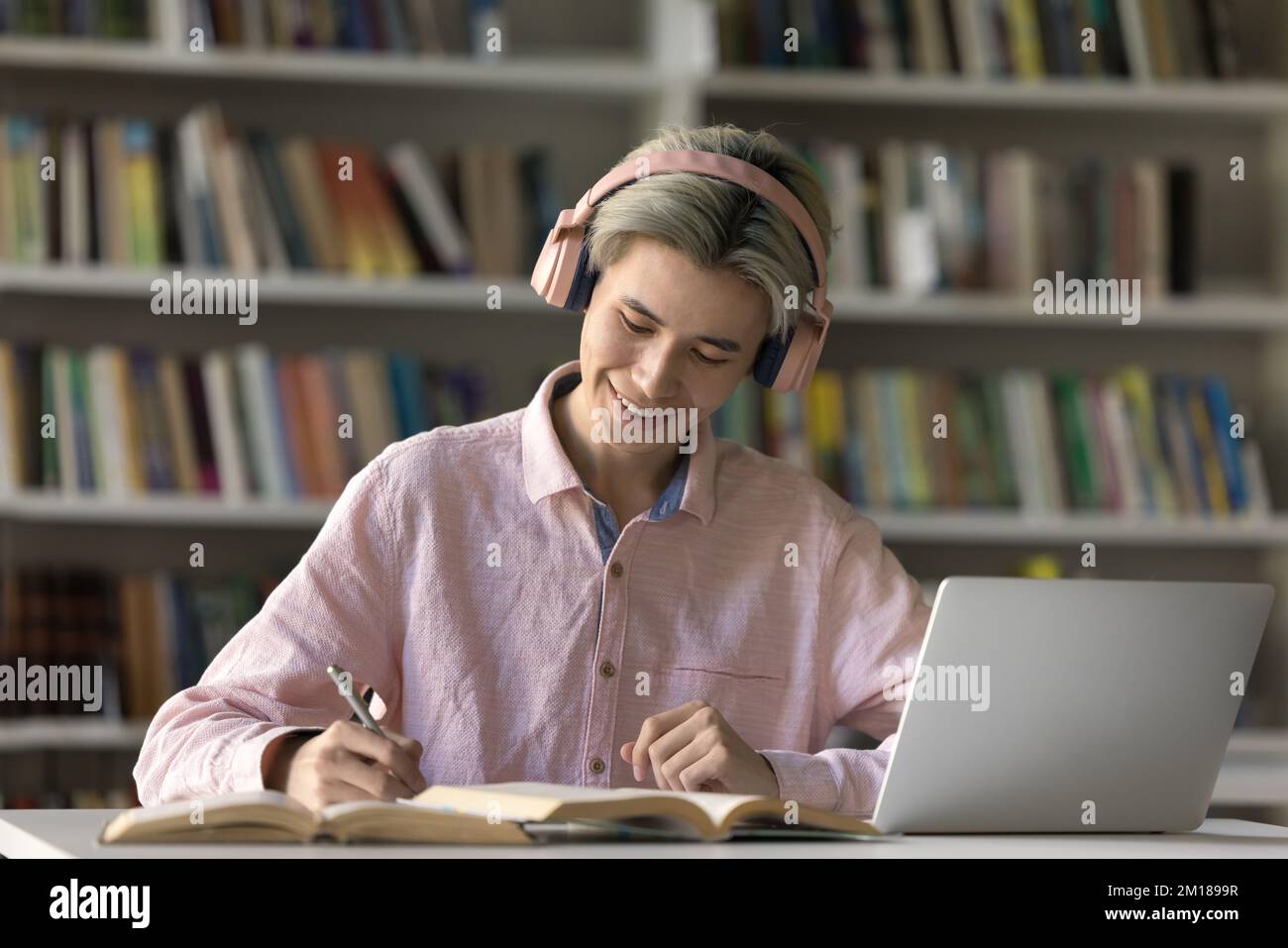 Focused student guy in pink wireless headphones studying in library ...