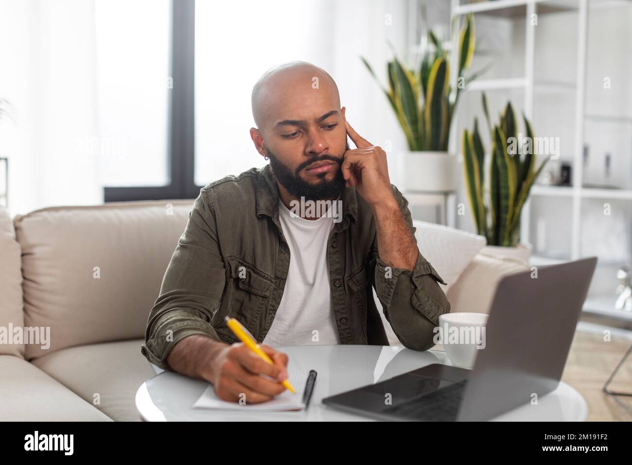 Serious man talking on cellphone and taking notes, looking at laptop ...