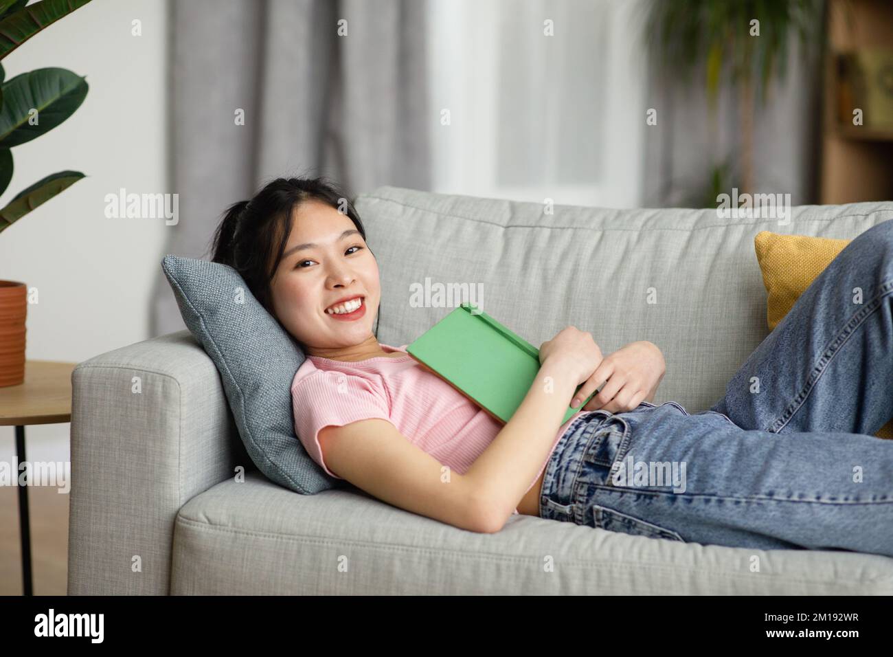 Stay home hobbies. Cheerful asian lady lying on sofa with book, looking ...