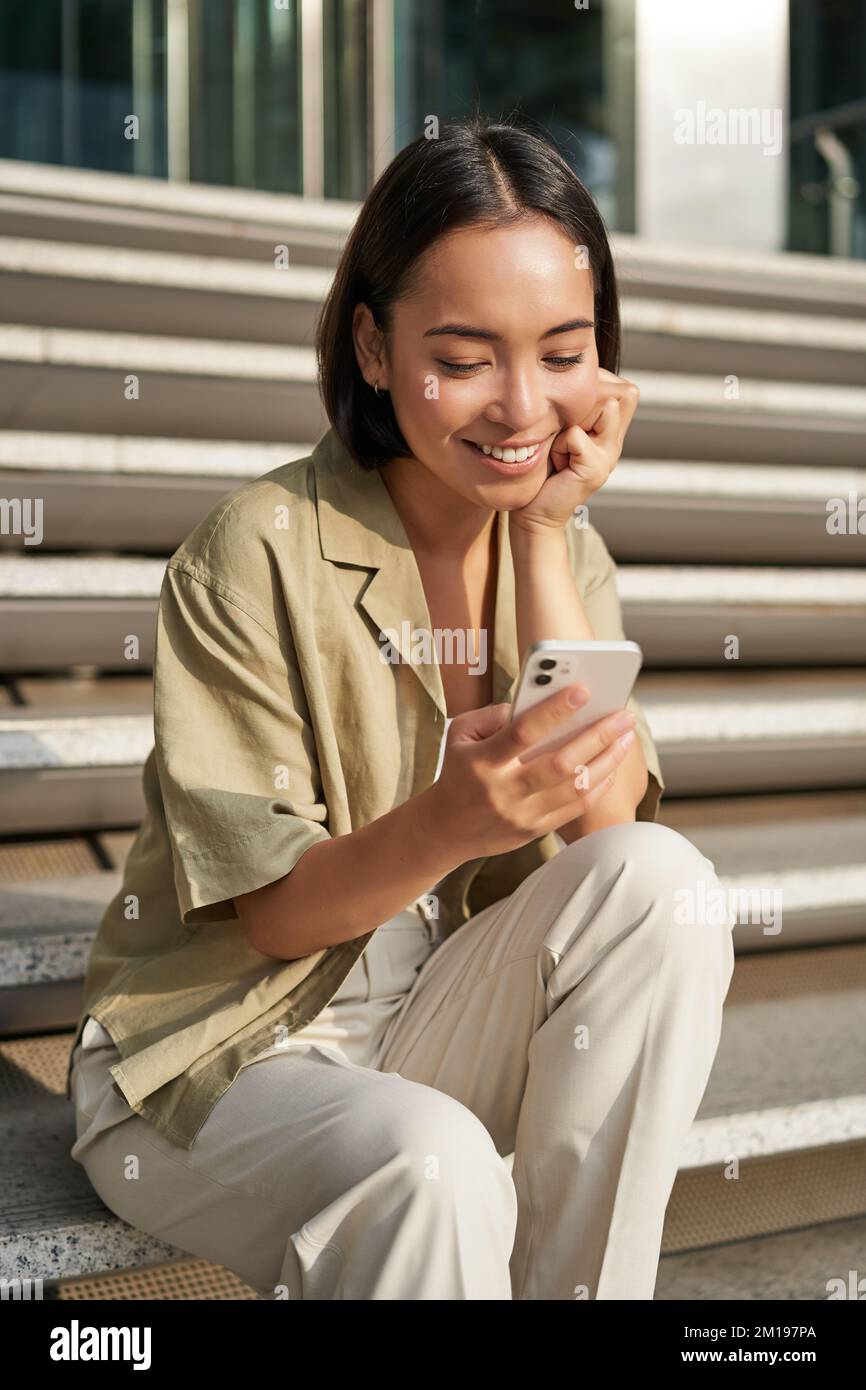 Vertical shot of asian woman, student sits on stairs in city, looking ...