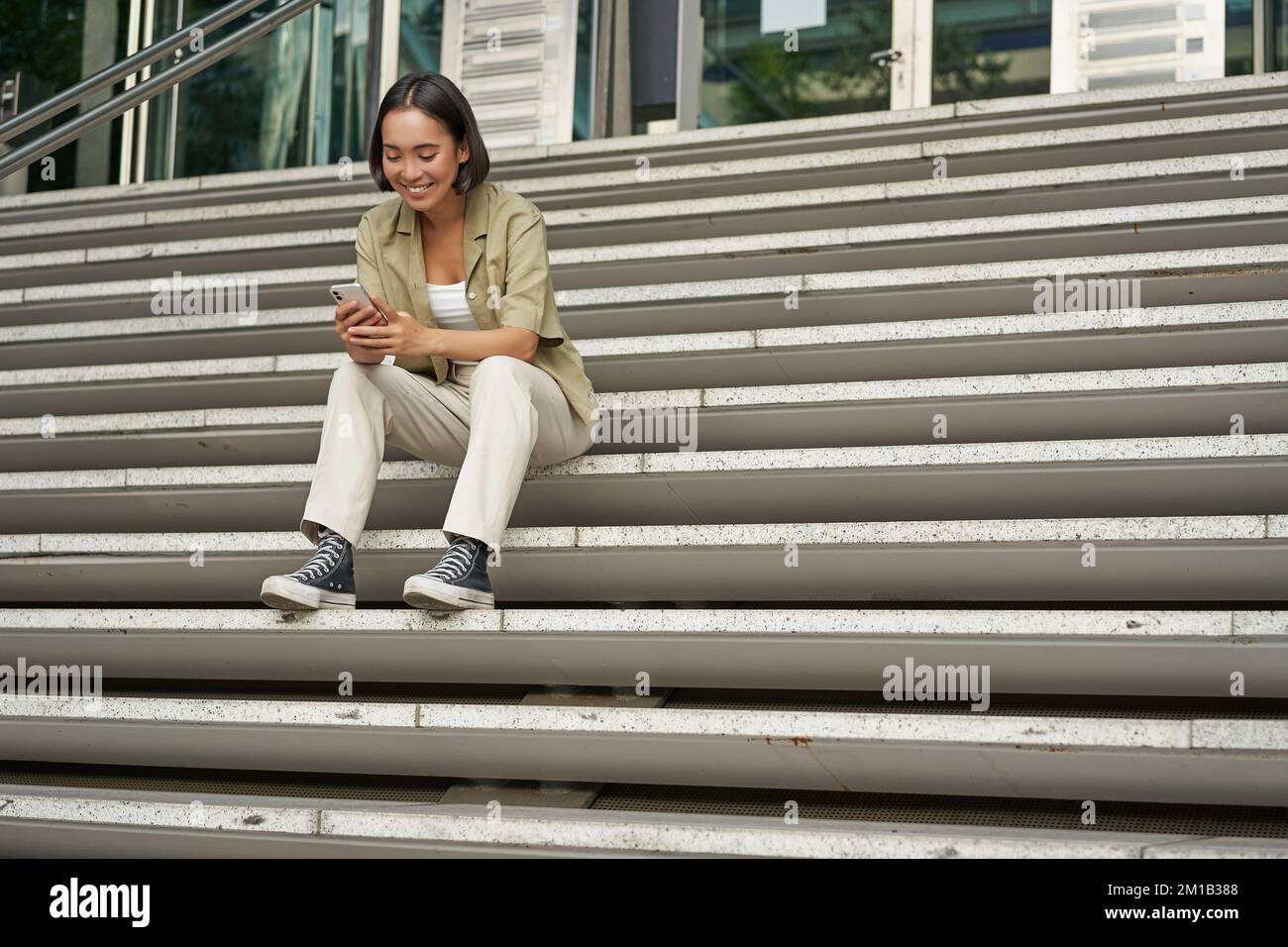 Portrait of smiling asian girl sits on stairs outdoors, sending message ...