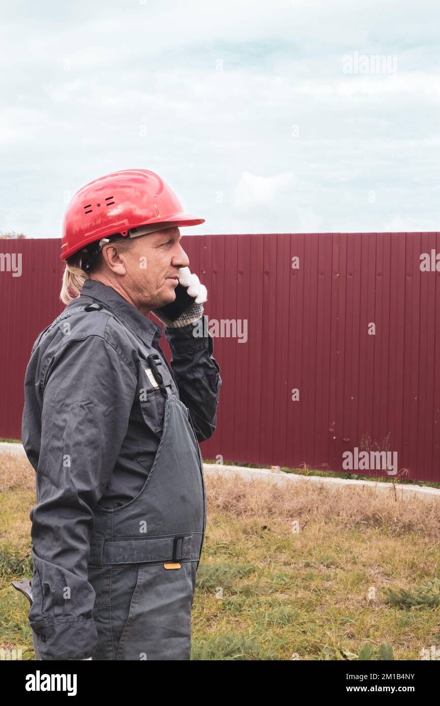 An adult male worker in overalls and a hard hat is talking on the phone ...