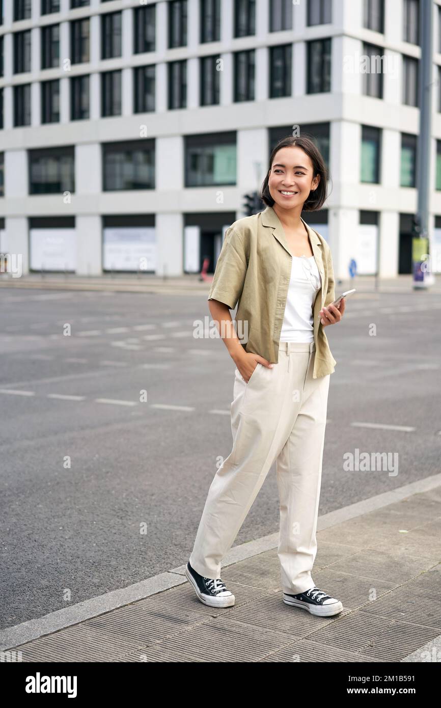 Woman smiling, standing with smartphone on street, waiting for taxi ...