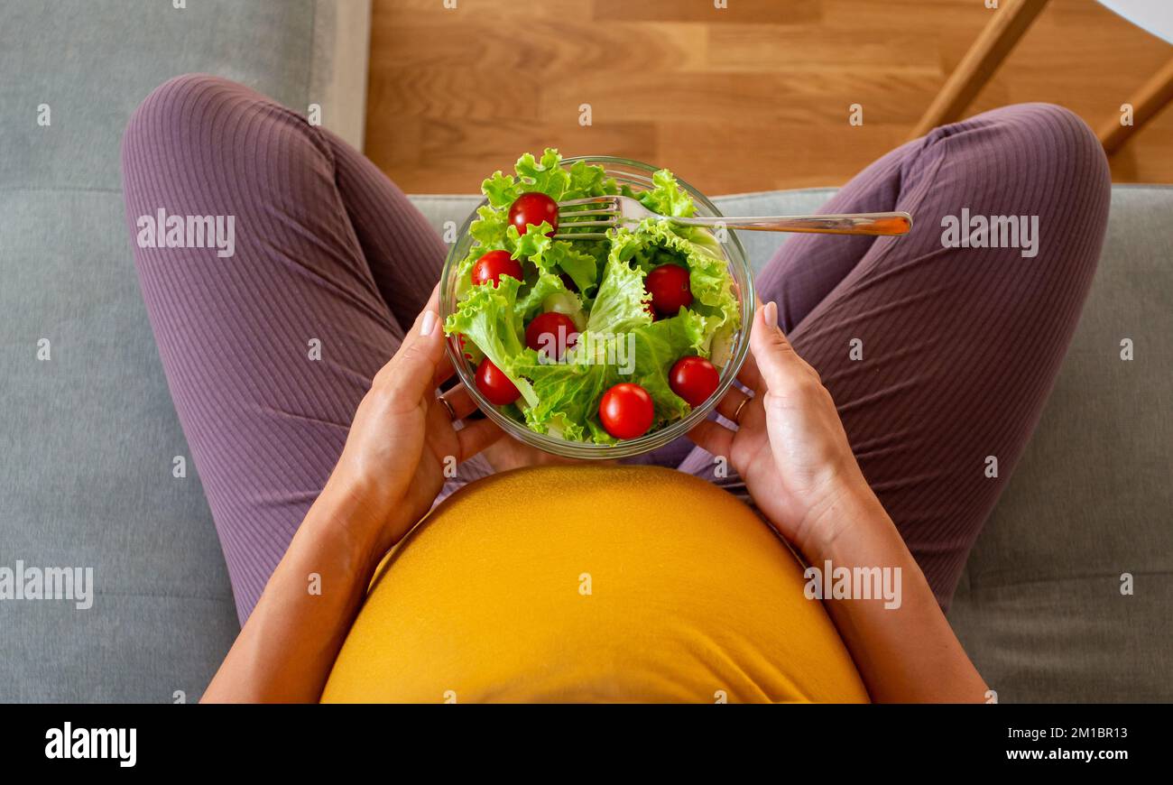 Pregnant woman sitting on sofa and holding bowl with salad, shoot ...