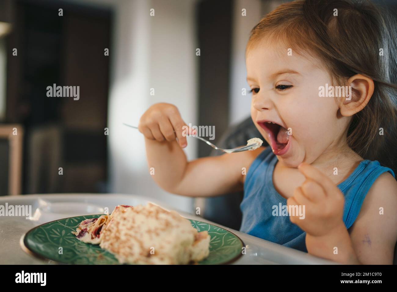 The baby girl in the kitchen at the table eating food with fork by ...