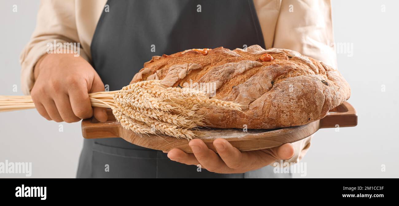 Man with fresh bread and wheat spikelets on light background, closeup ...