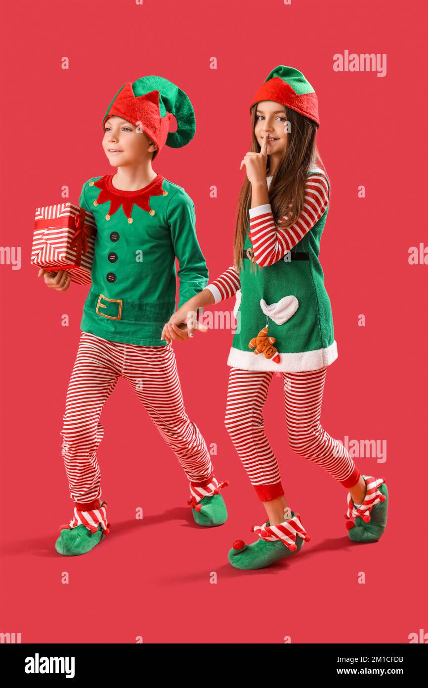 Little children in elf pajamas with Christmas gift on red background ...