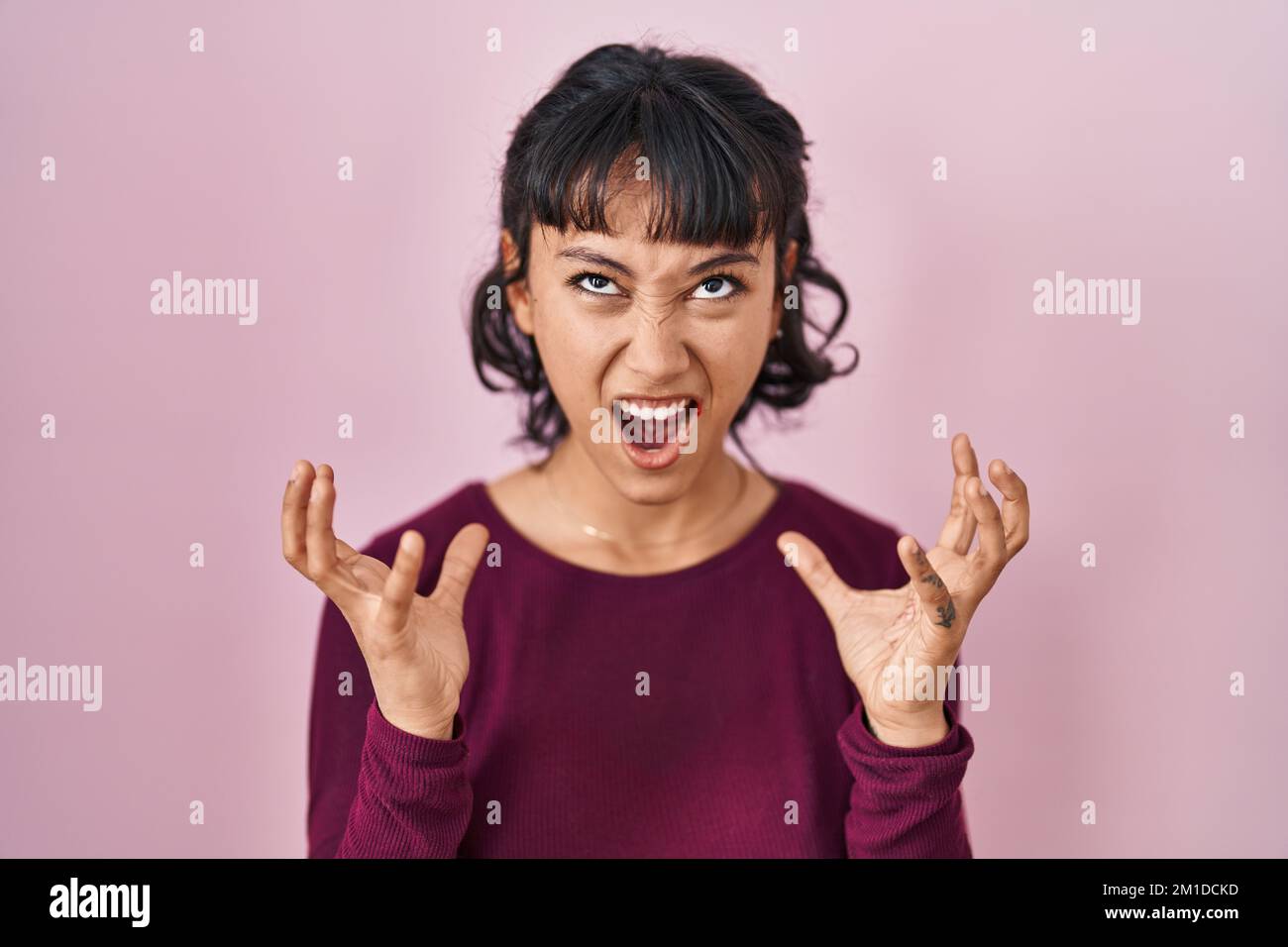 Young beautiful woman standing over pink background crazy and mad ...