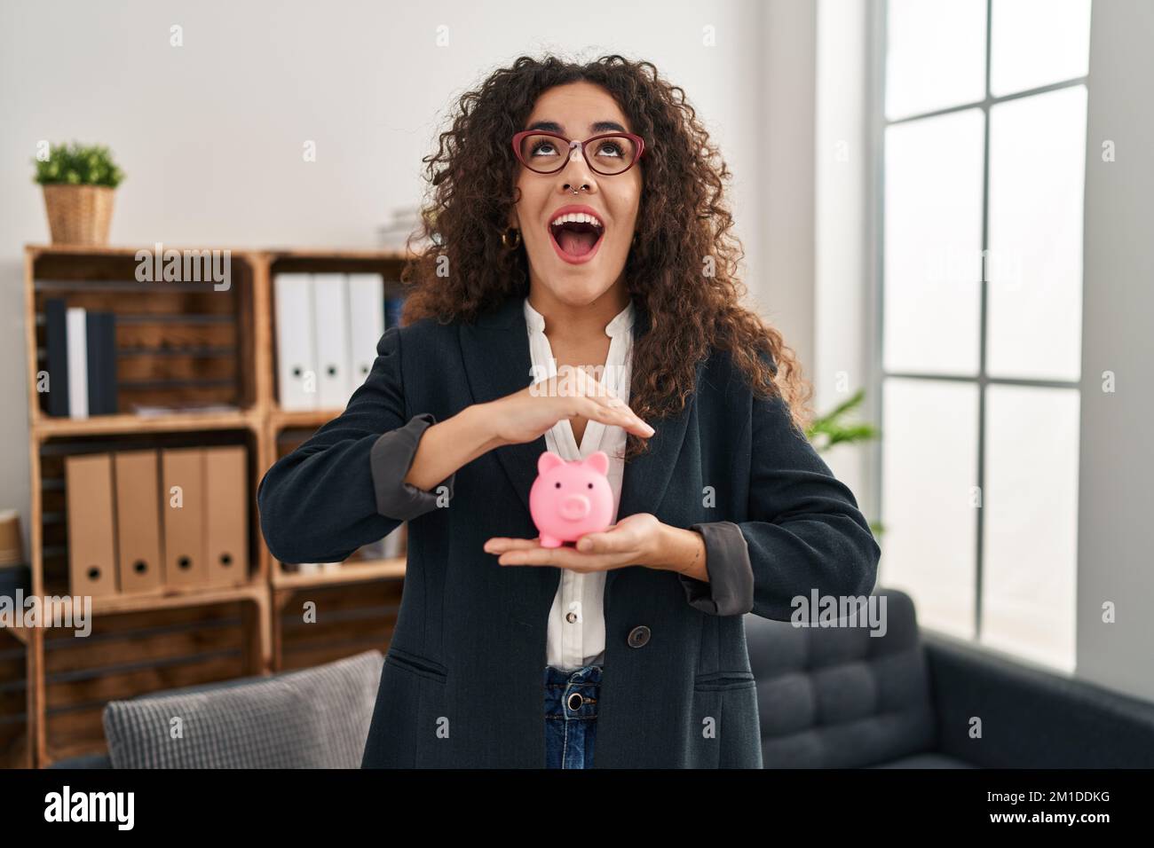 Young hispanic woman holding piggy bank angry and mad screaming ...