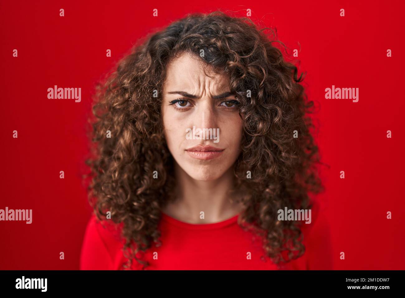 Hispanic woman with curly hair standing over red background skeptic and ...