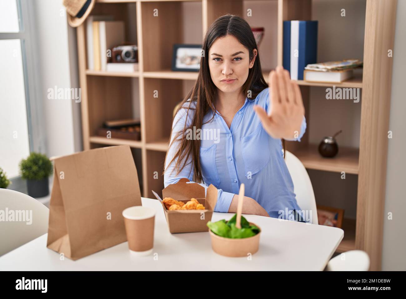 Young brunette woman eating take away food at home with open hand doing ...
