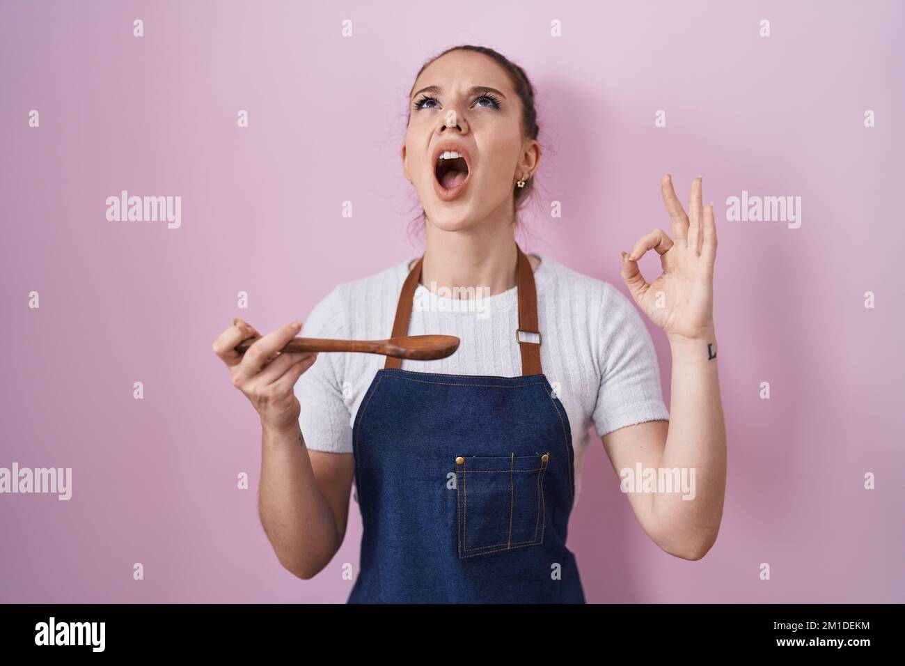 Young hispanic girl wearing professional cook apron holding wood spoon ...