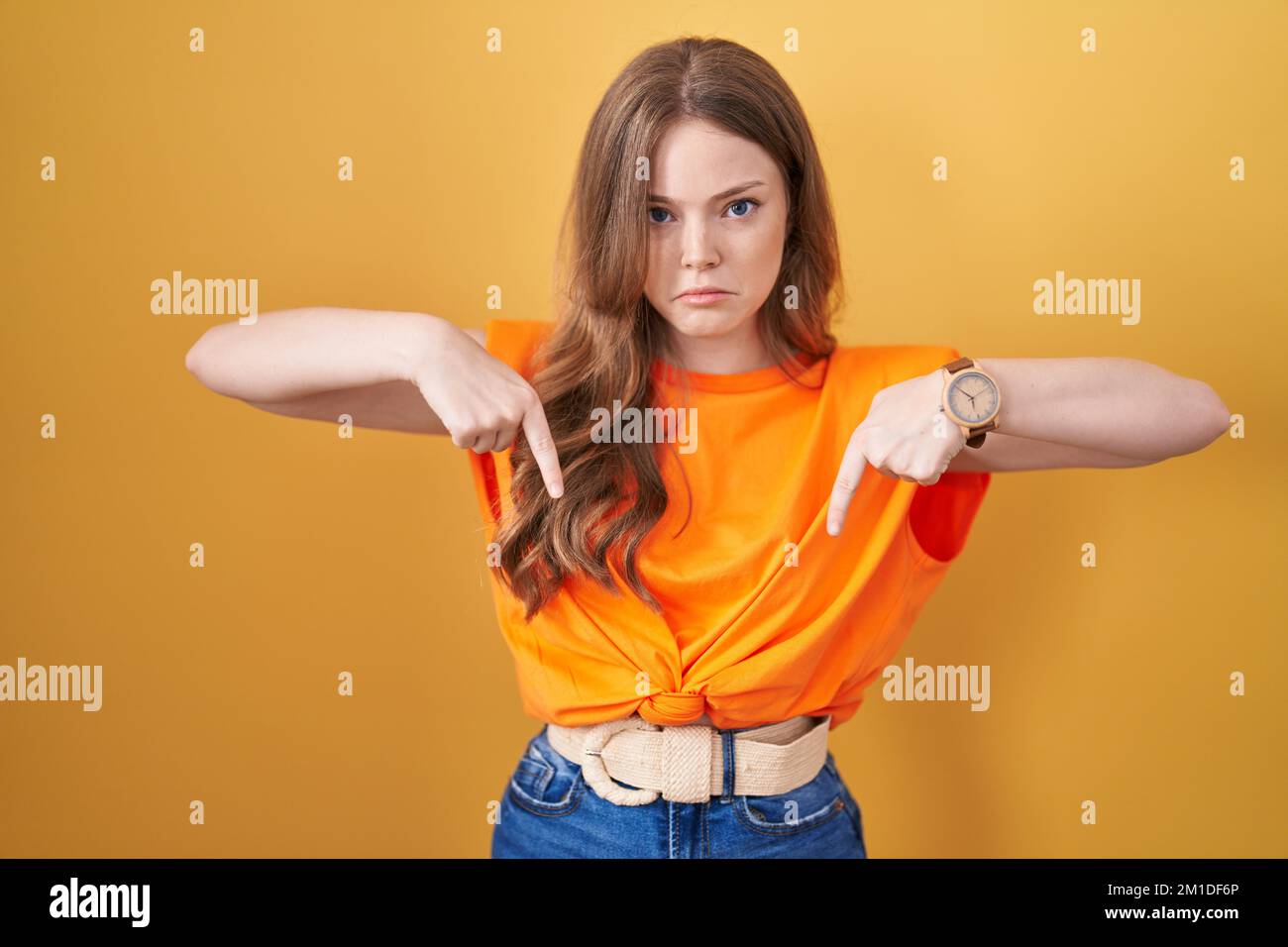 Caucasian woman standing over yellow background pointing down looking ...