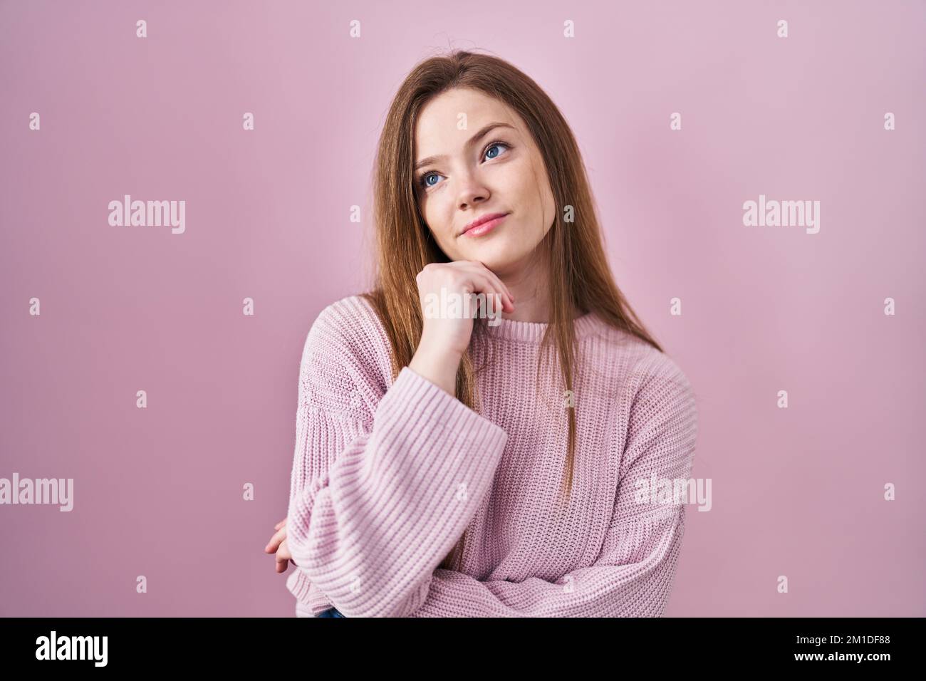 Young caucasian woman standing over pink background with hand on chin ...