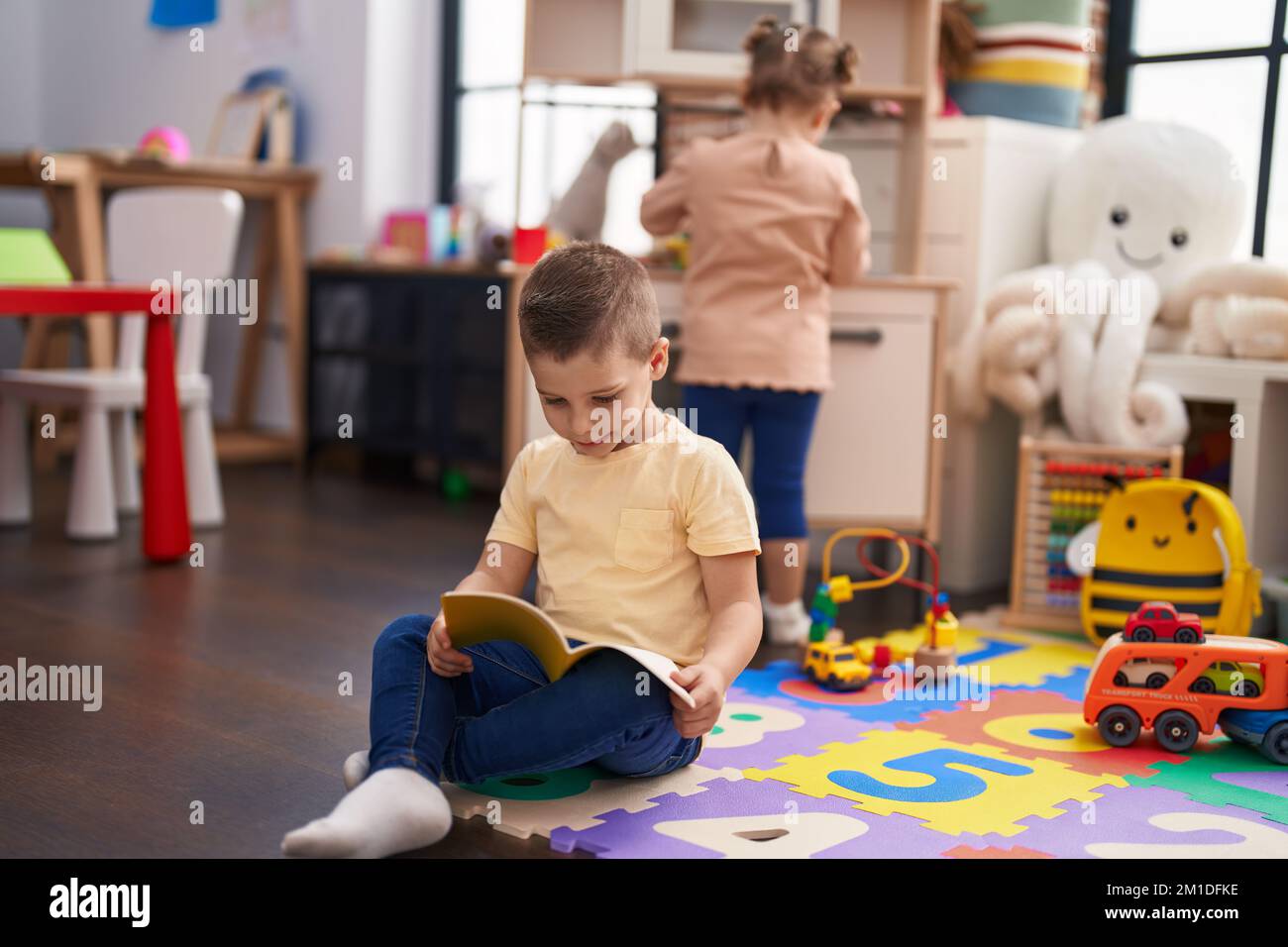 Adorable toddler reading book sitting on floor at kindergarten Stock ...