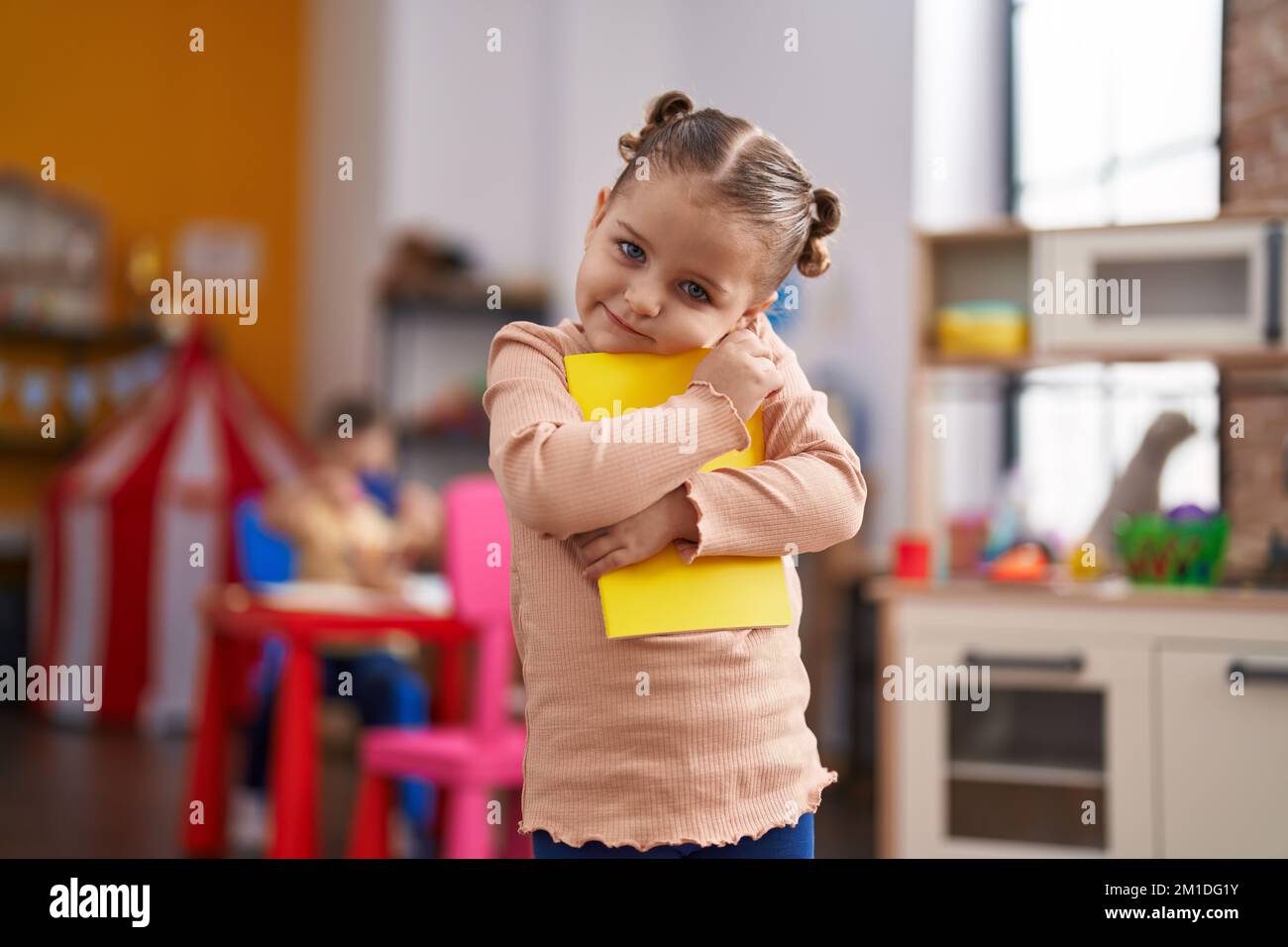 Adorable hispanic girl student hugging book standing at kindergarten ...