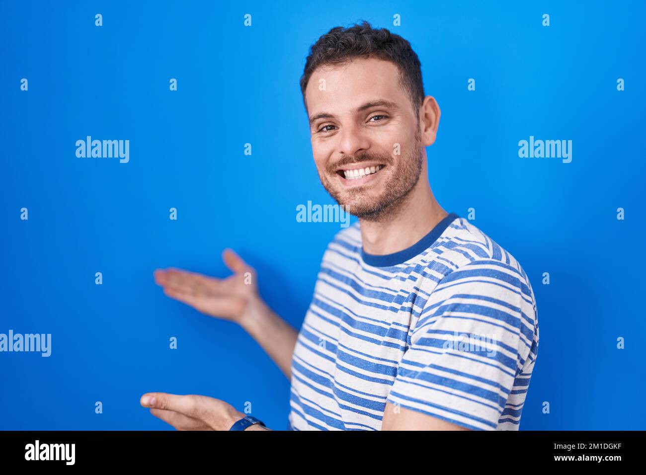 Young hispanic man standing over blue background inviting to enter ...