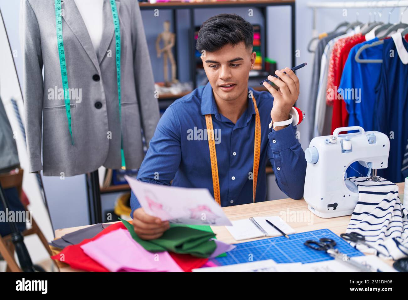 Young hispanic man tailor talking on smartphone looking clothing design ...