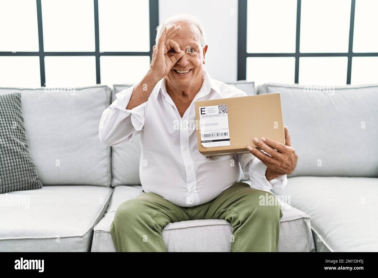 Senior man holding delivery package at home smiling happy doing ok sign ...