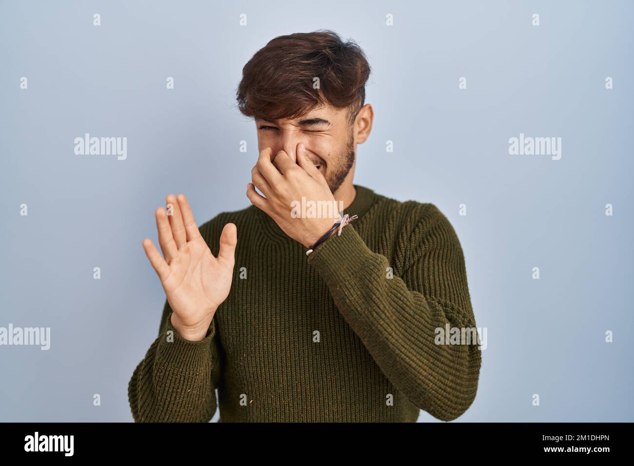 Arab man with beard standing over blue background smelling something ...
