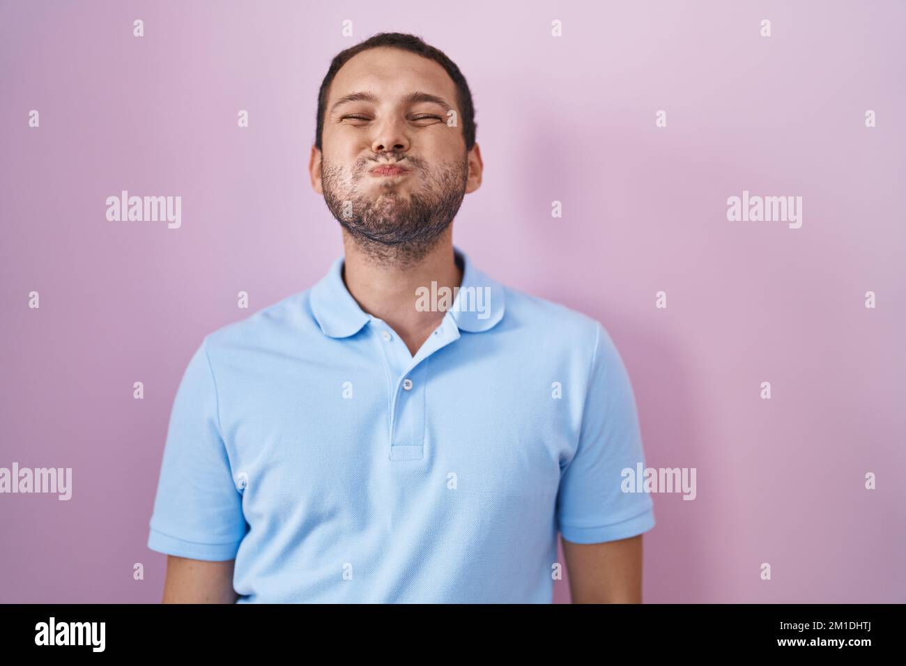Hispanic man standing over pink background puffing cheeks with funny ...