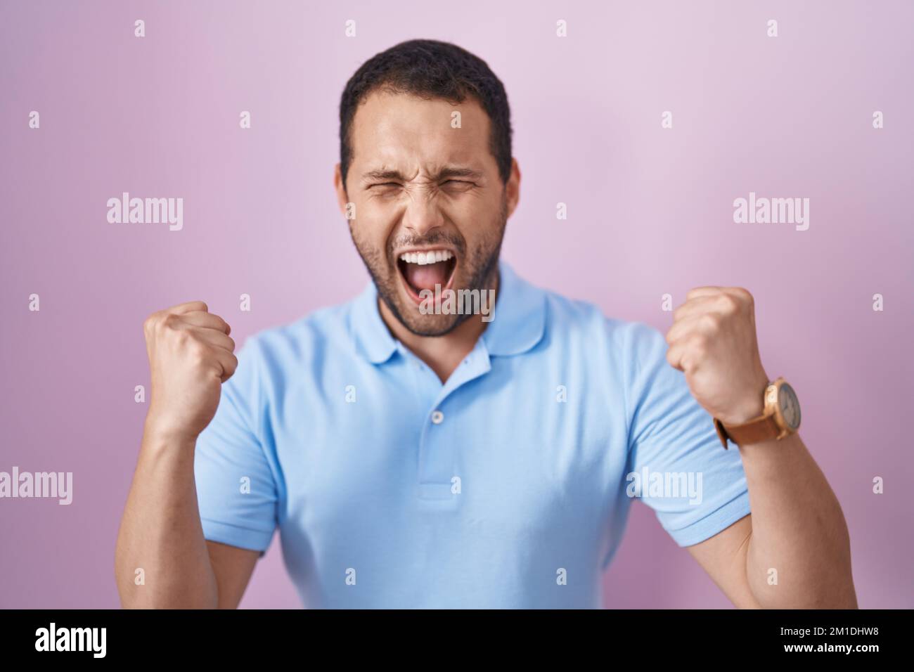 Hispanic man standing over pink background angry and mad raising fists ...