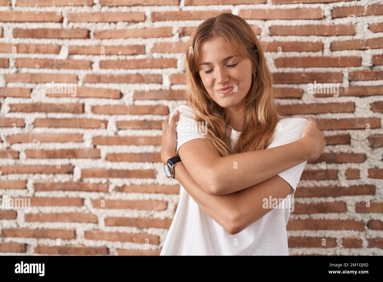 Young caucasian woman standing over bricks wall hugging oneself happy ...