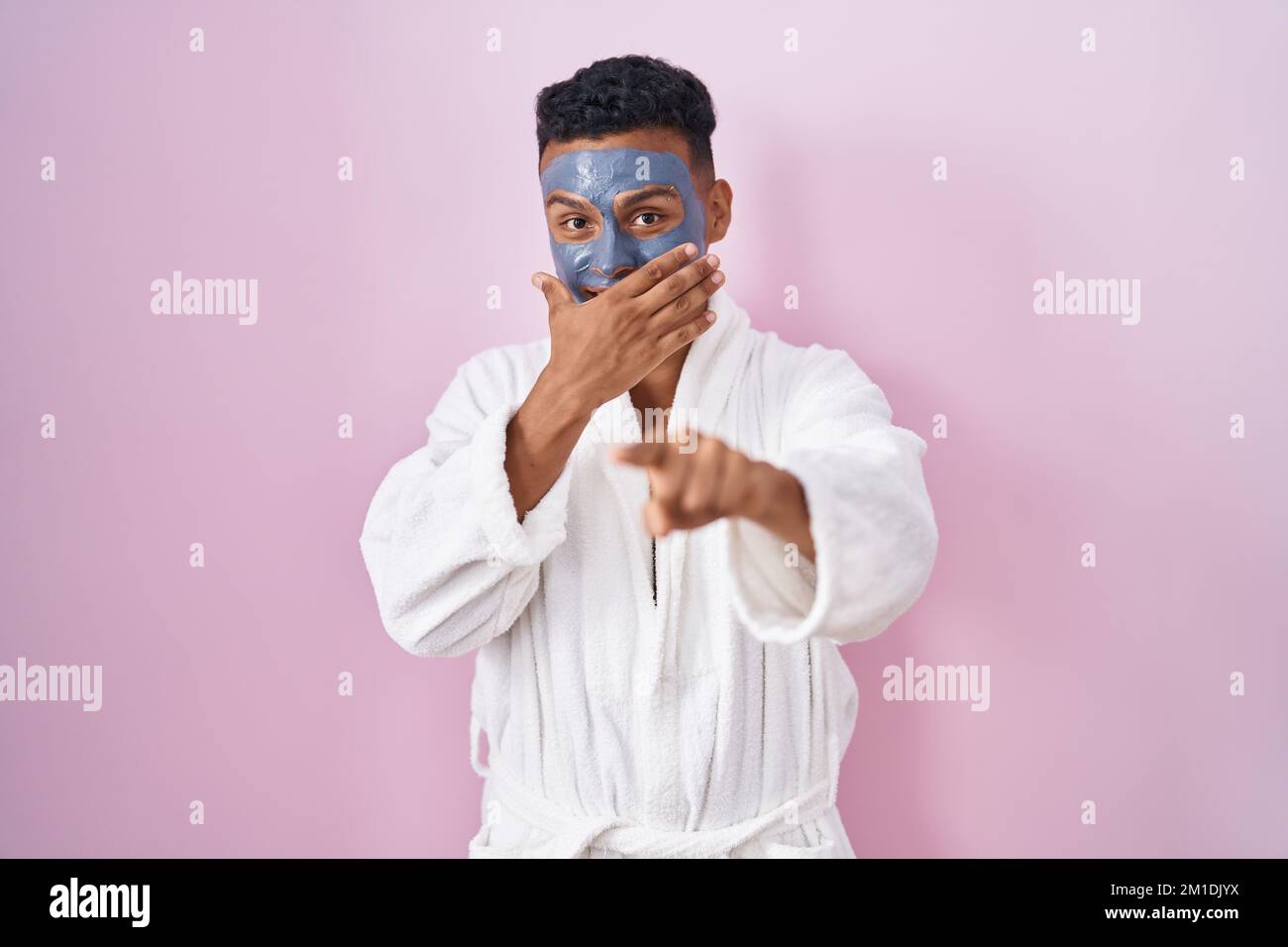 Young hispanic man wearing beauty face mask and bath robe laughing at ...