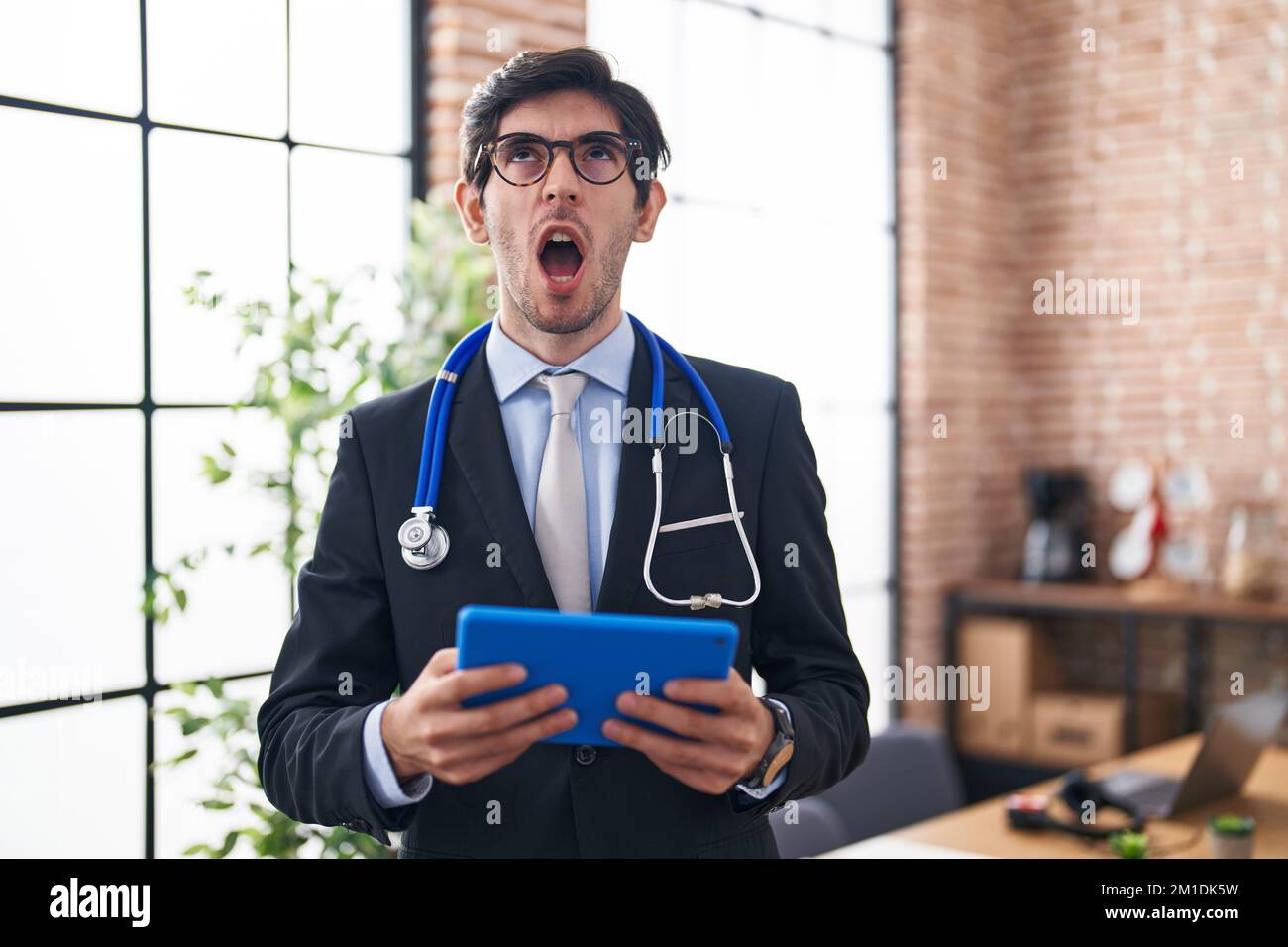Young hispanic man wearing doctor stethoscope at the office angry and ...