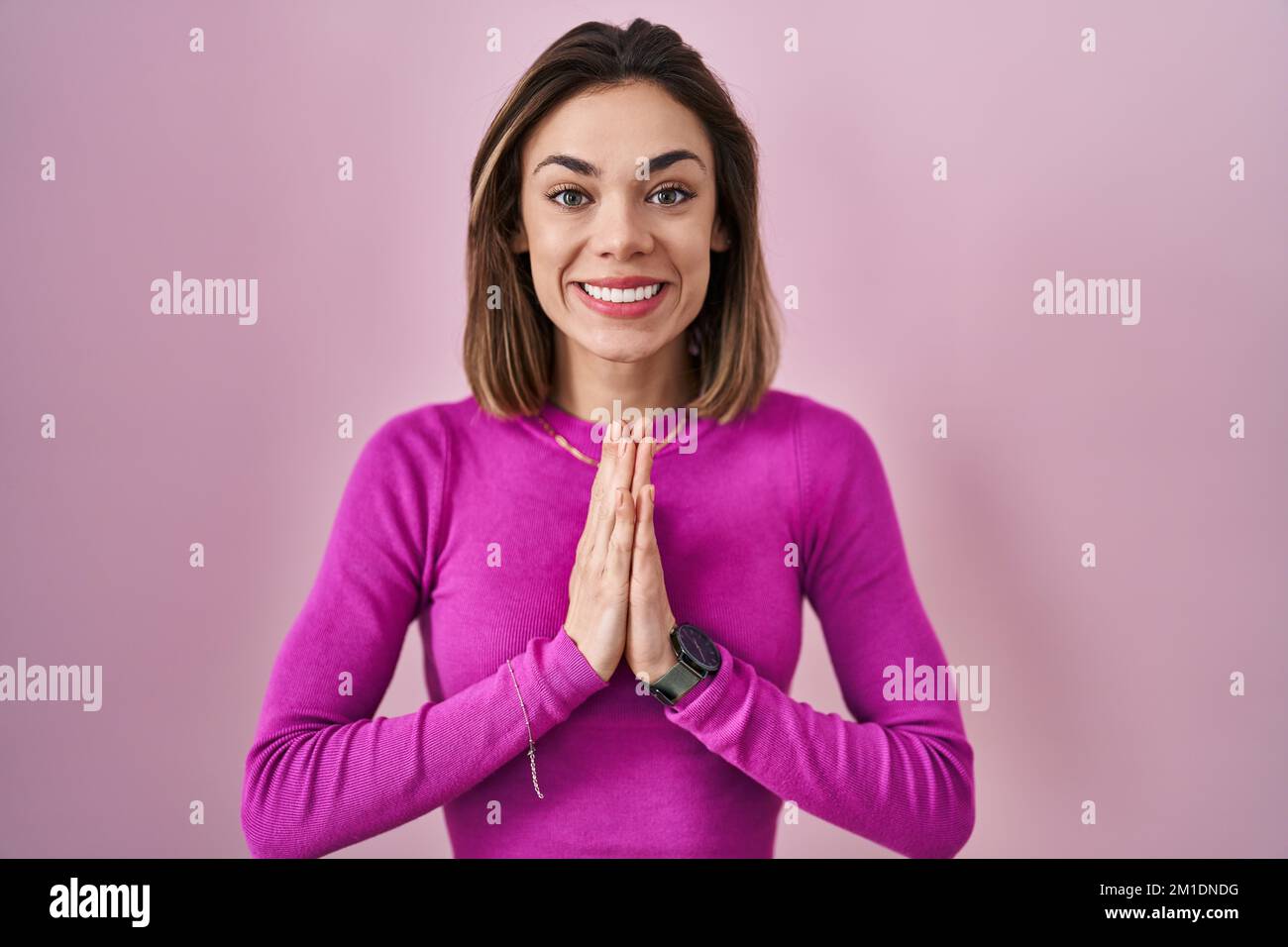 Hispanic woman standing over pink background praying with hands ...