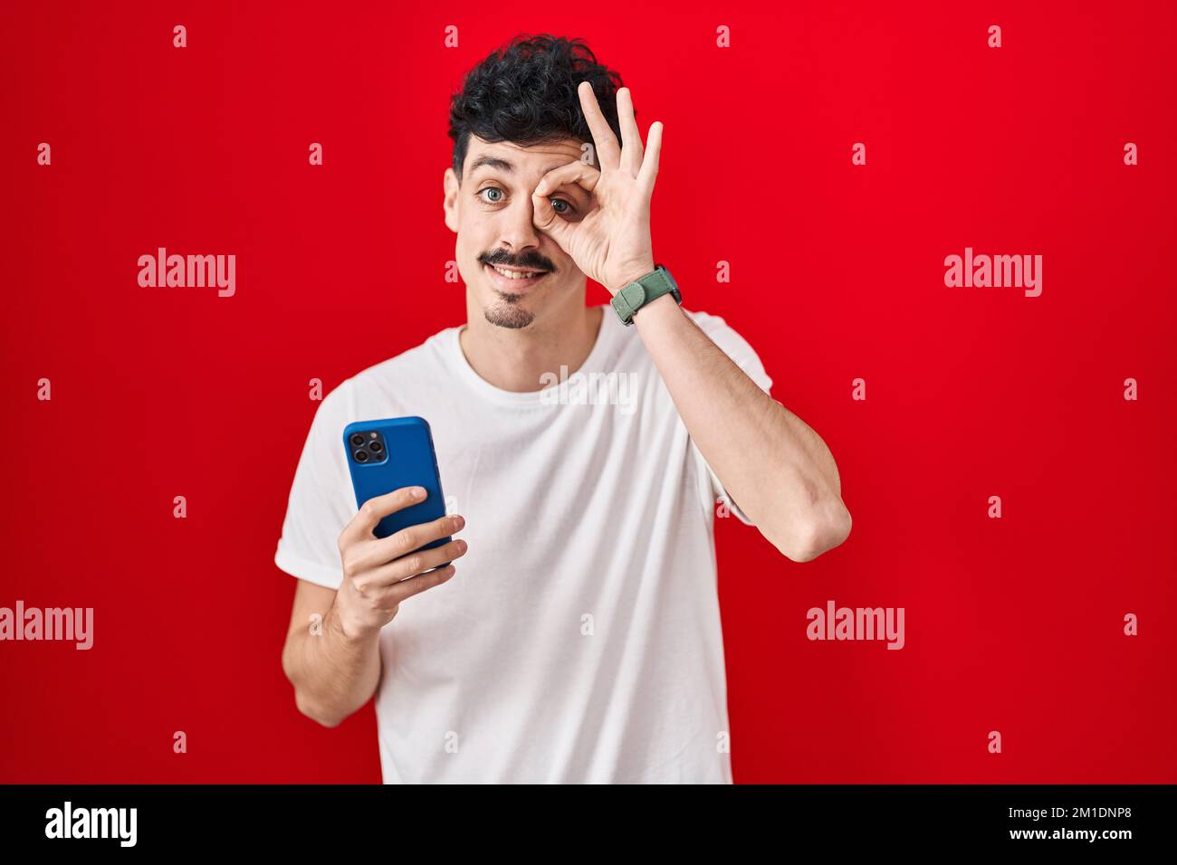 Hispanic man using smartphone over red background doing ok gesture with ...