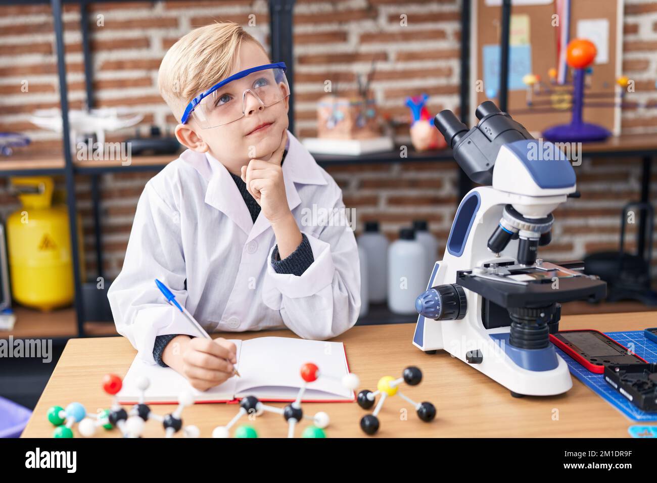 Adorable toddler student writing on notebook with doubt expression at ...