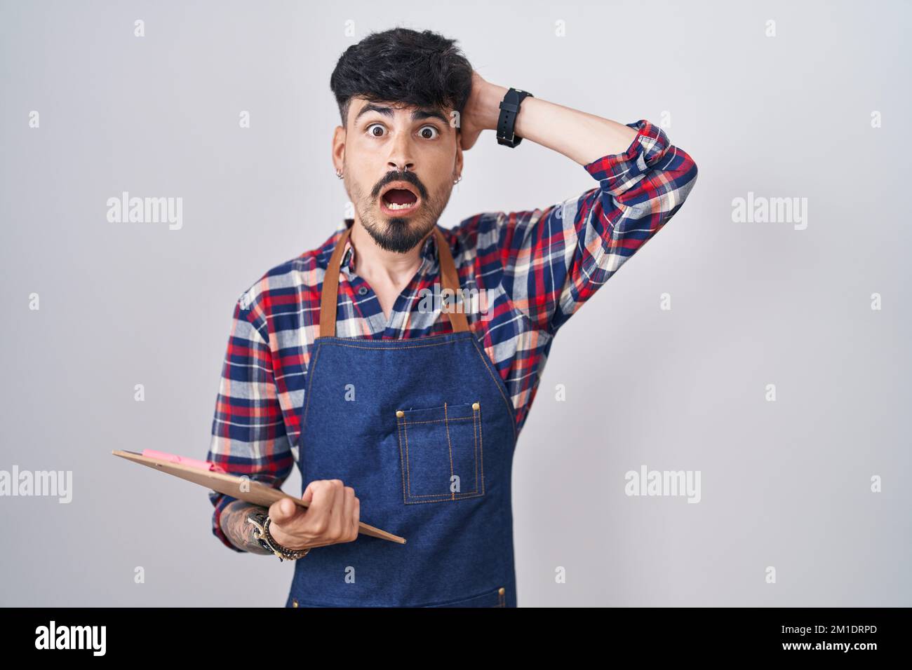 Young hispanic man with beard wearing waiter apron holding clipboard ...