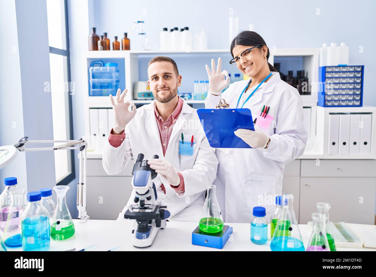 Two young people working at scientist laboratory doing ok sign with ...