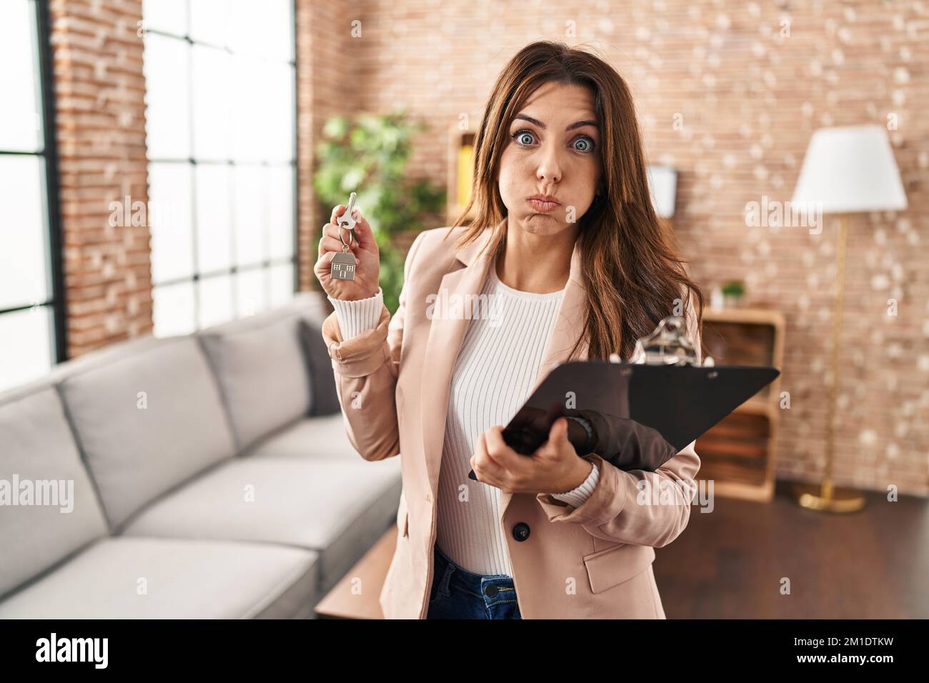 Young brunette woman working as real state agent holding keys of new home puffing cheeks with ...