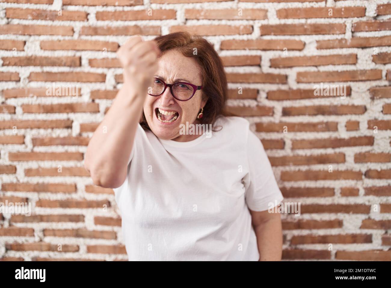 Senior woman with glasses standing over bricks wall angry and mad ...