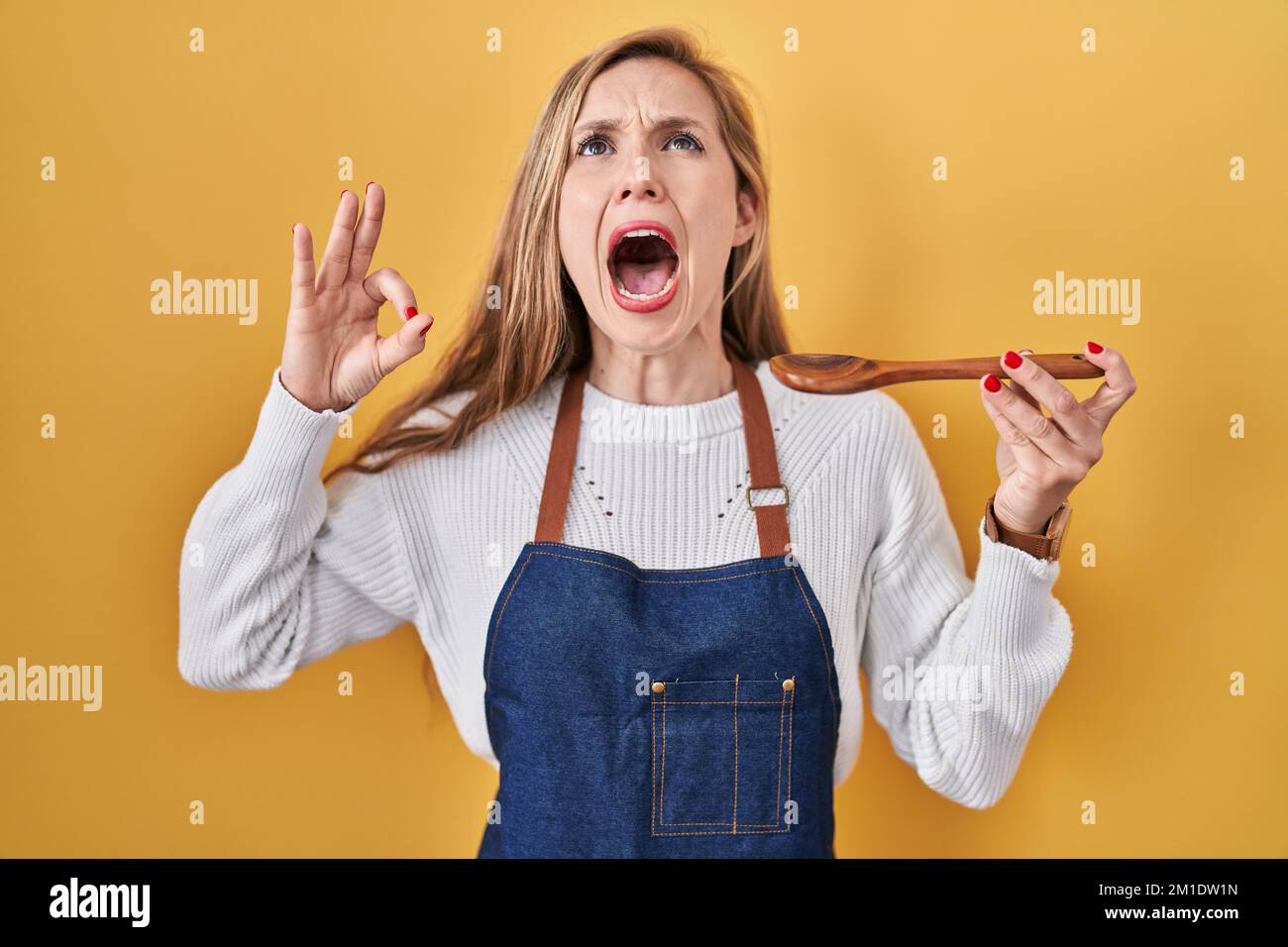 Young blonde woman wearing apron tasting food holding wooden spoon ...
