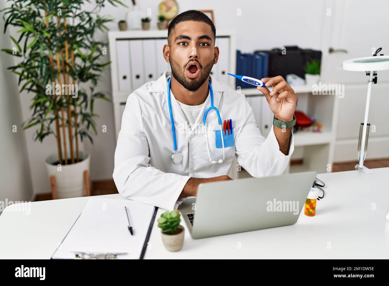 Young indian doctor holding thermometer scared and amazed with open ...