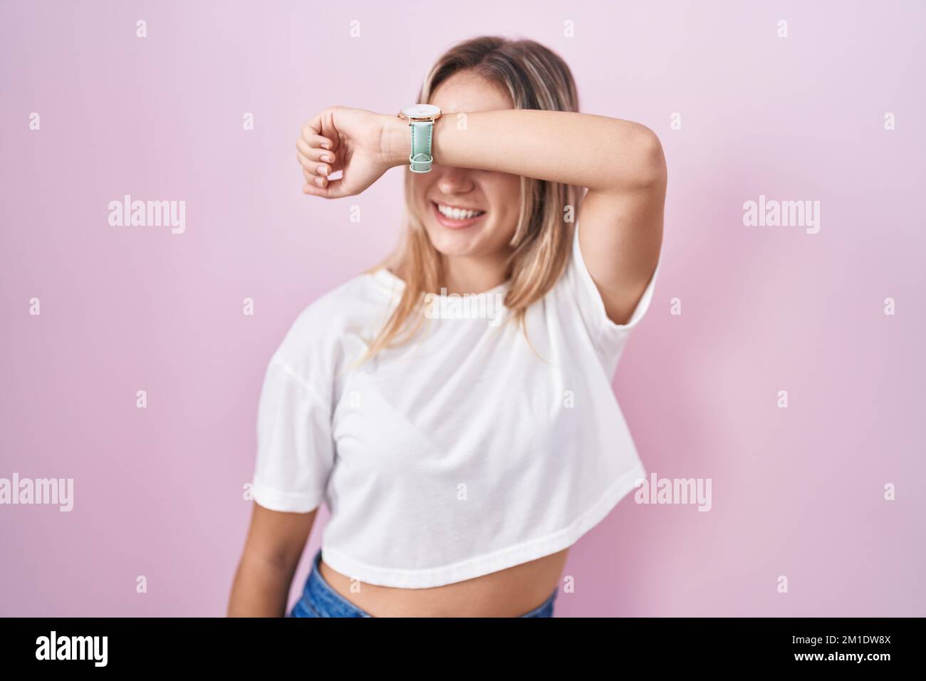 Young blonde woman standing over pink background covering eyes with arm ...